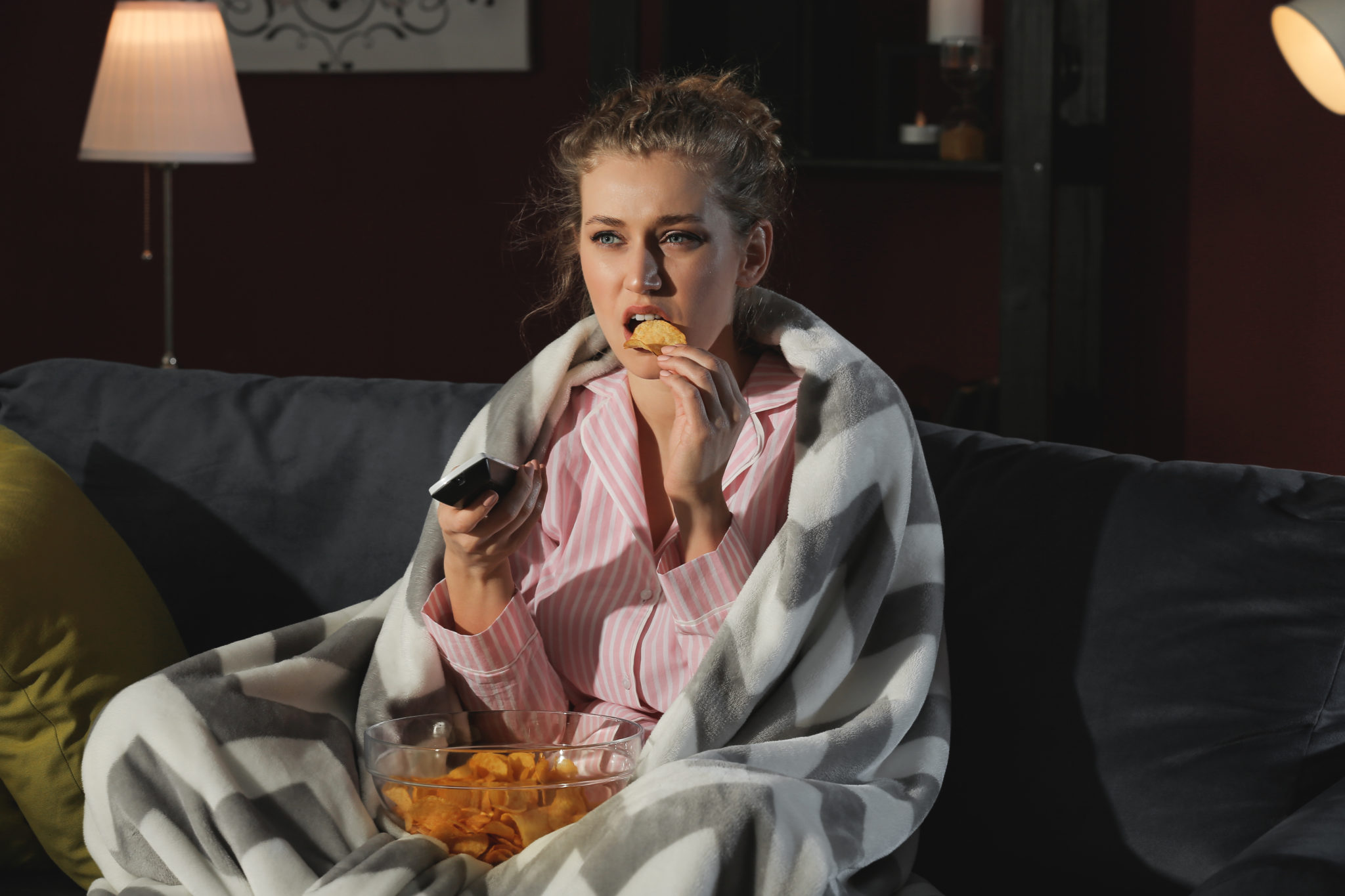 Beautiful young woman eating unhealthy food while watching TV at night ...